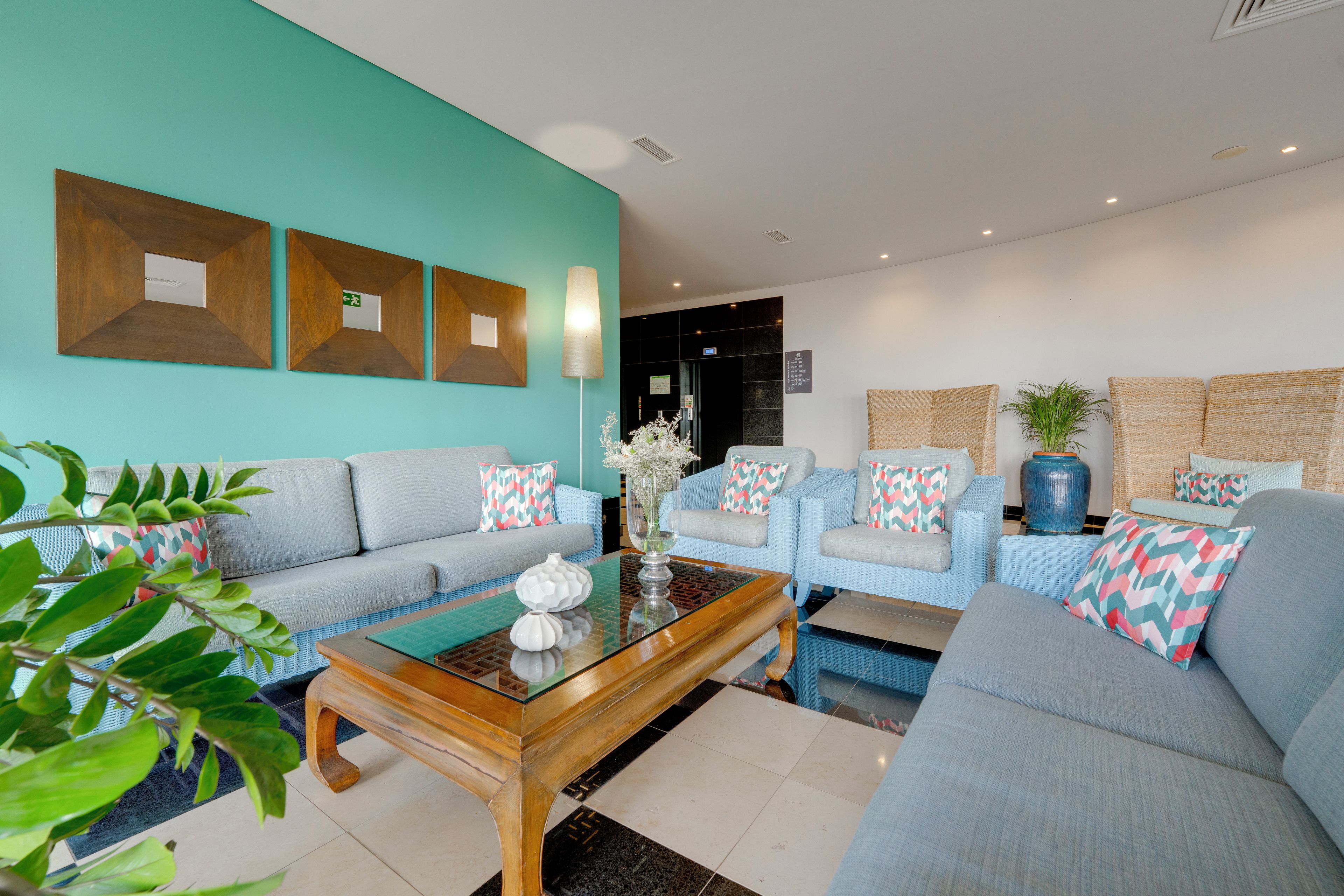 Um hall de hotel moderno com uma parede azul-esverdeada, uma mesa de centro de madeira e dois sofás com almofadas estampadas.