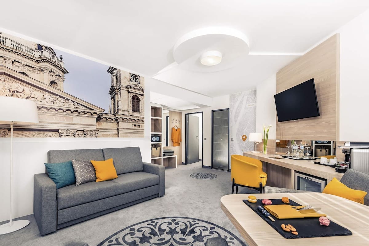 Quarto de hotel moderno com sofá, mesa de jantar, televisão e vista para um edifício histórico.