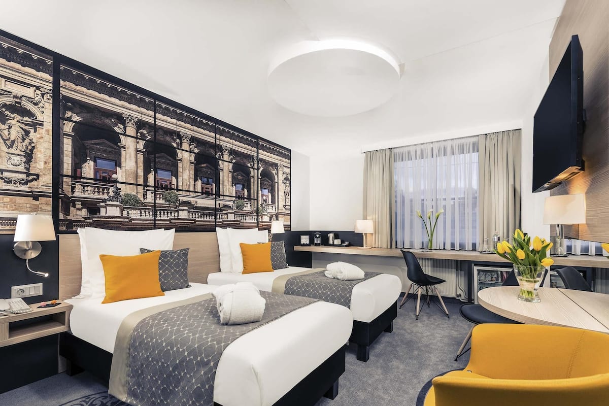 Quarto de hotel com duas camas, uma escrivaninha, uma cadeira e um grande painel decorativo.
