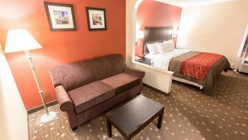 Quarto de hotel com sofá marrom, uma mesinha, cama com cobertor vermelho e duas molduras na parede.