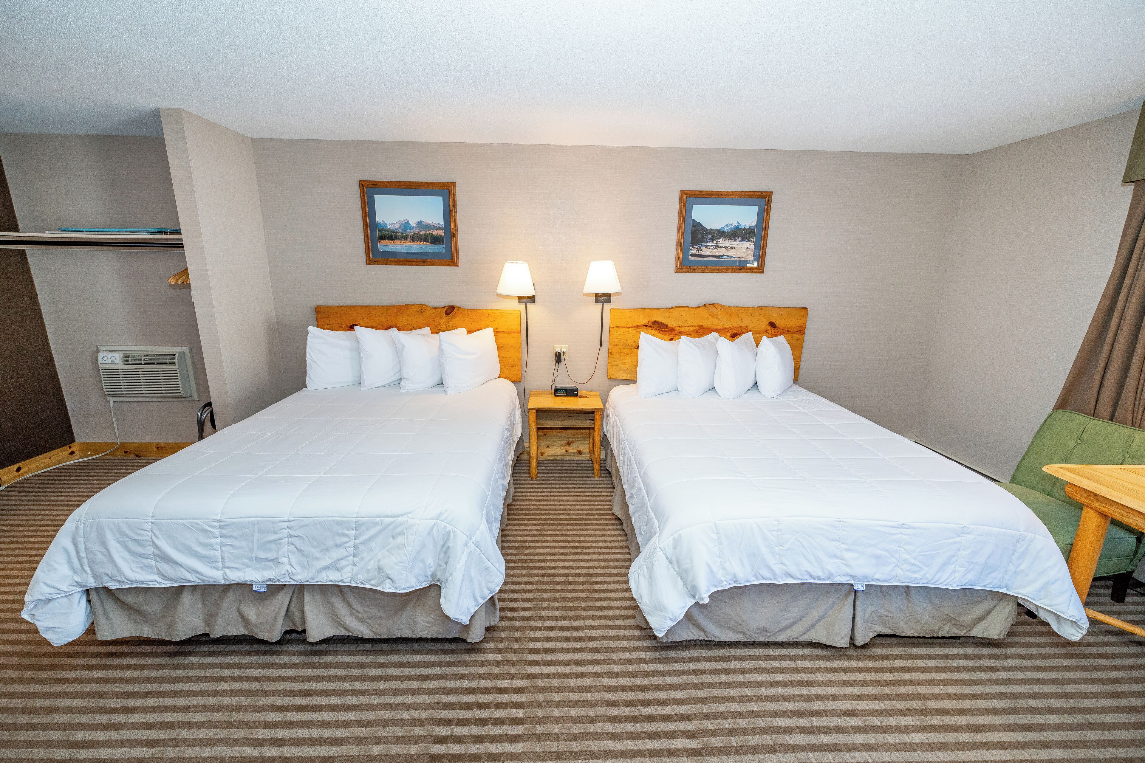 Quarto de hotel com duas camas, uma cabeceira de madeira, mesa de cabeceira com abajur, uma cadeira verde e uma pequena mesa.