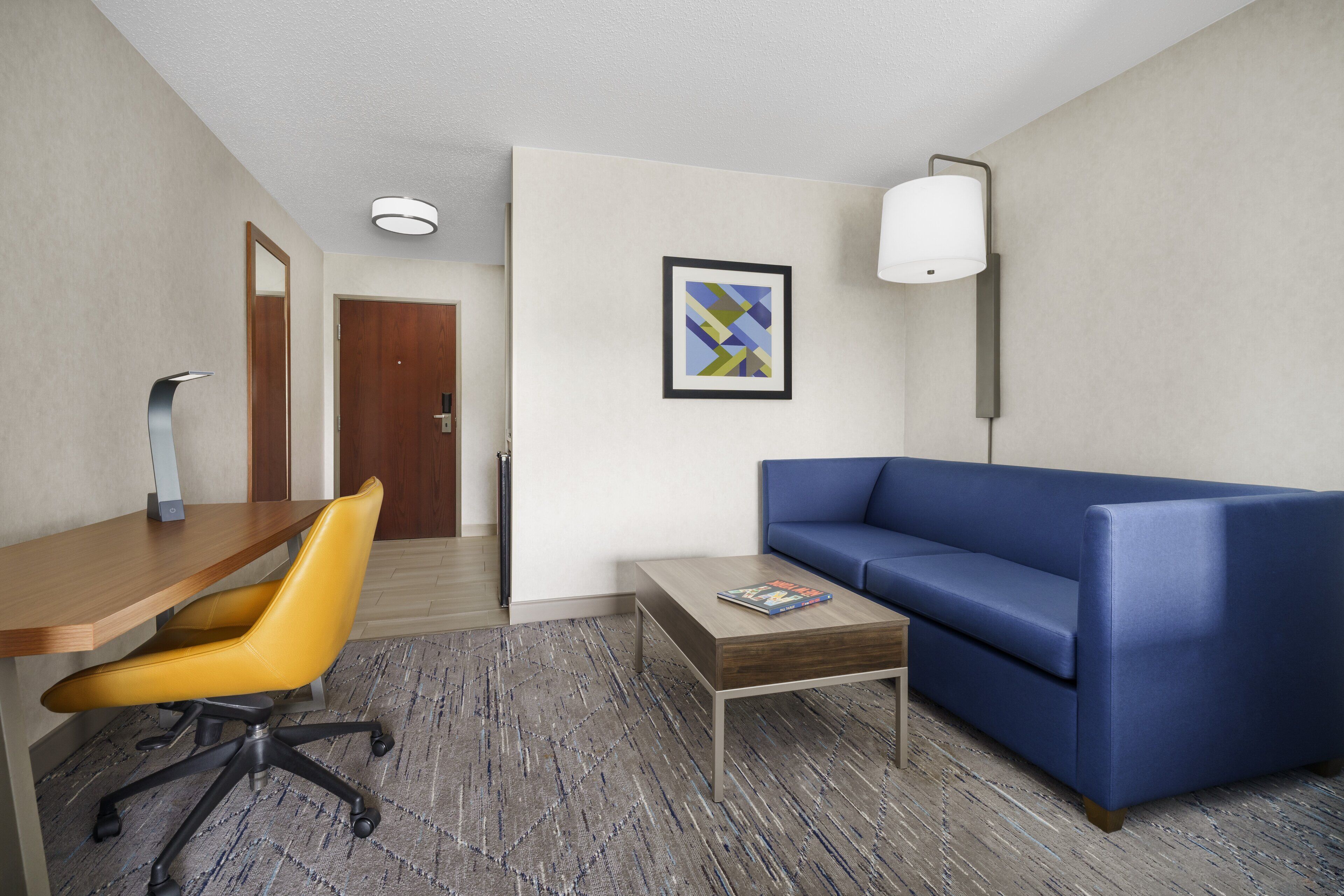 Quarto de hotel moderno com escrivaninha de madeira, cadeira de escritório amarela, sofá azul, mesinha de centro e uma pintura abstrata emoldurada na parede.