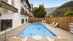 Piscina com espreguiçadeiras e uma mesa, cercada por um muro de madeira e um edifício com varandas.