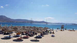Uma praia com espreguiçadeiras e guarda-sóis, uma torre de salva-vidas e veleiros ao fundo.
