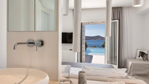 Quarto de hotel moderno com uma pia grande branca, televisão de tela plana, cama com roupa de cama branca e vista para uma praia e oceano.