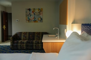 Quarto de hotel com cama, pequeno espaço de cozinha, sofá e uma obra de arte na parede.