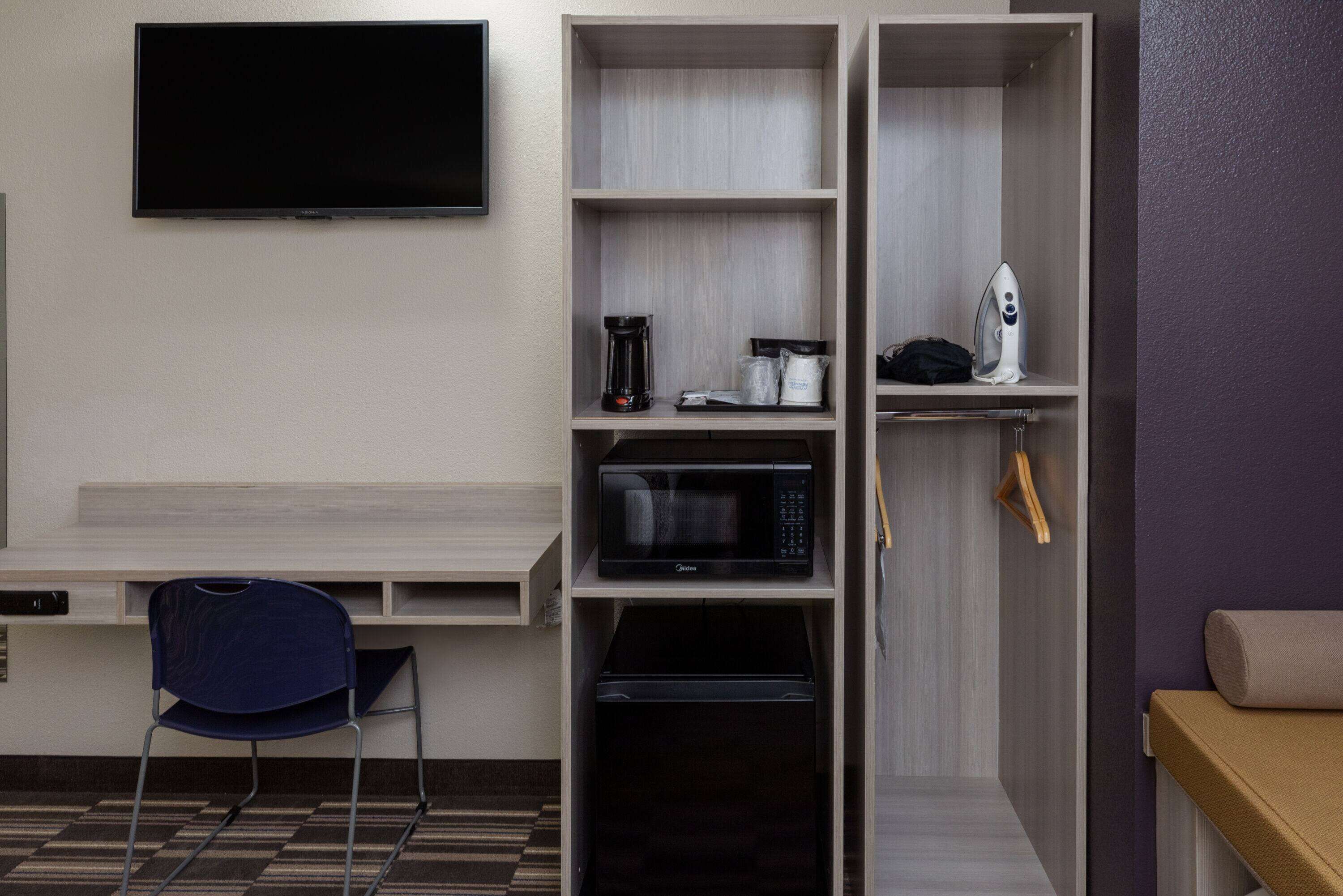 Quarto de hotel com televisão de tela plana na parede, micro-ondas, cafeteira e tábua de passar.