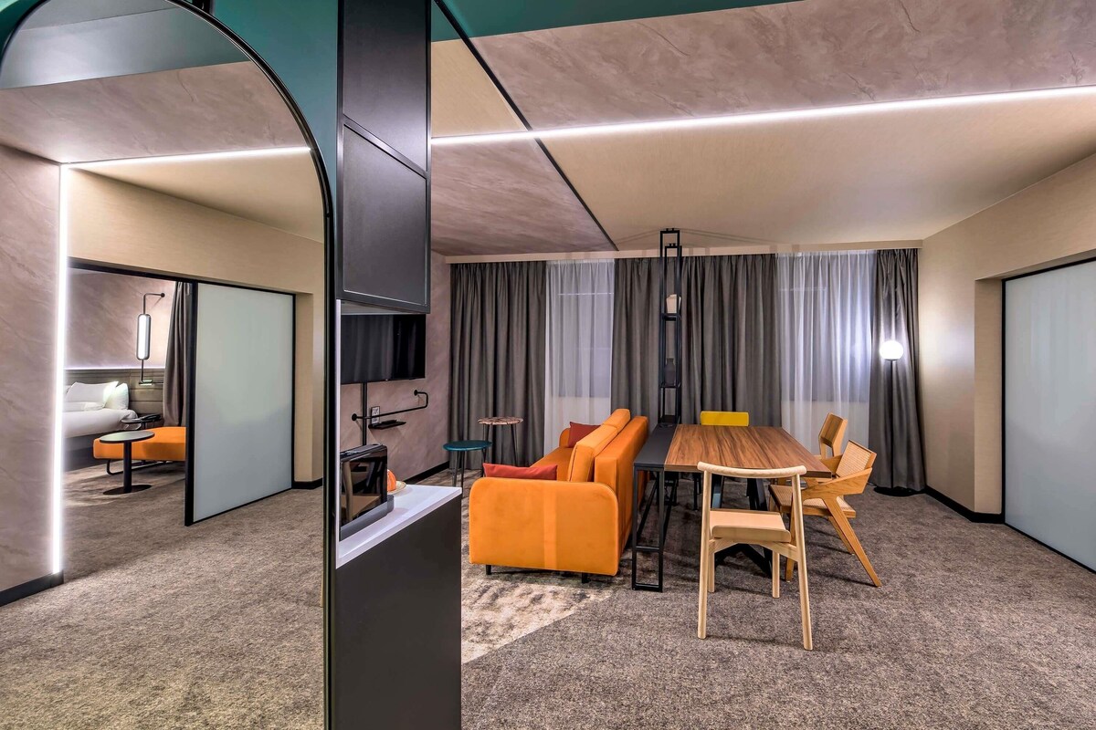 Quarto de hotel moderno com uma cama grande, uma escrivaninha, uma cadeira, uma mesa de jantar com cadeiras e uma janela ampla com cortinas.