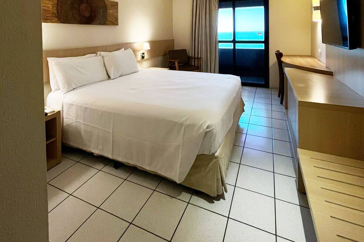 Quarto de hotel com uma cama grande, uma escrivaninha, uma televisão e vista para o mar pela janela.