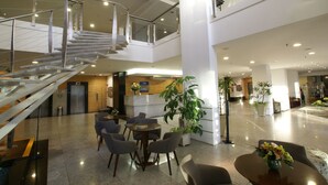 Um hall amplo com escadaria central, área de estar, balcão de recepção e plantas em vasos.