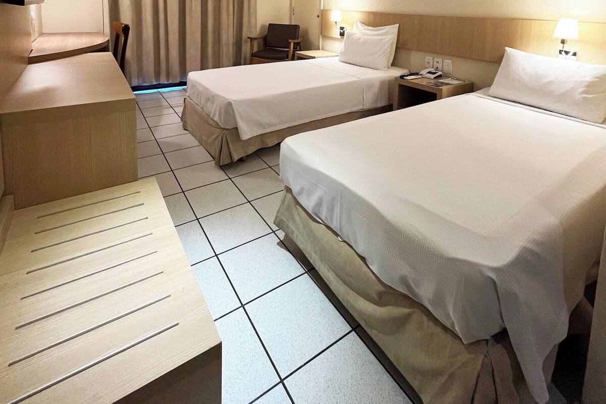 Quarto de hotel com duas camas, uma escrivaninha, uma cadeira, televisão e um quadro na parede.