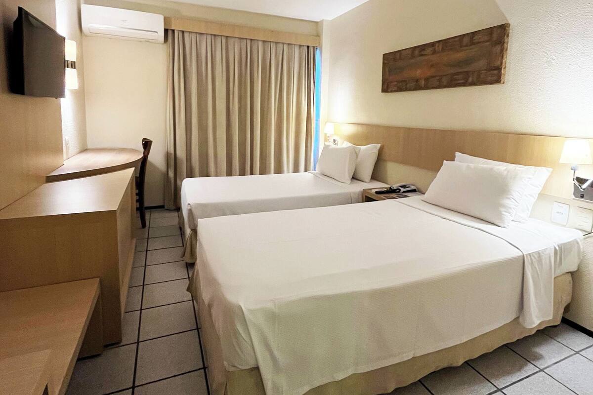 Quarto de hotel com duas camas, uma escrivaninha, uma televisão e uma janela com cortinas.