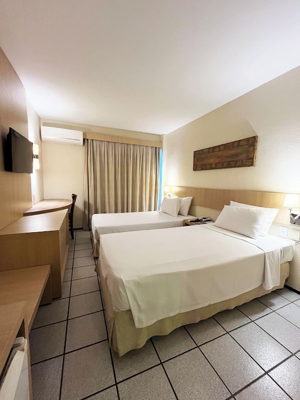 Quarto de hotel com duas camas, uma escrivaninha, uma televisão e uma janela com cortinas.