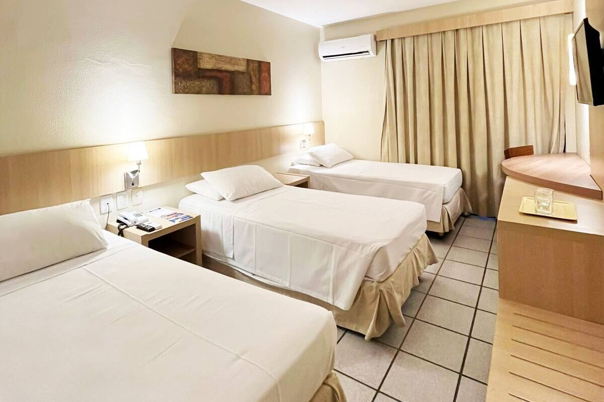 Quarto de hotel com três camas, uma escrivaninha, uma cadeira, uma televisão e um aparelho de ar condicionado.