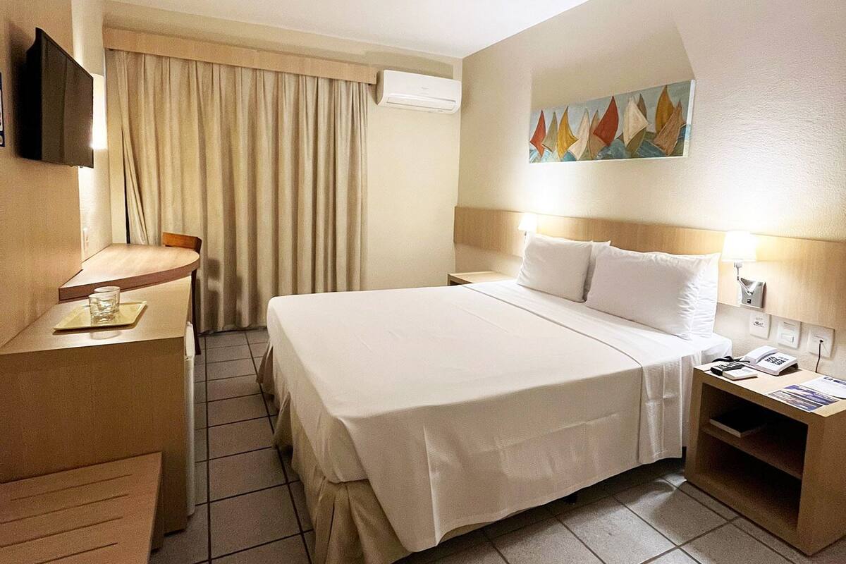 Quarto de hotel com cama, mesa de cabeceira, luminária, aparelho de ar condicionado e um quadro na parede.