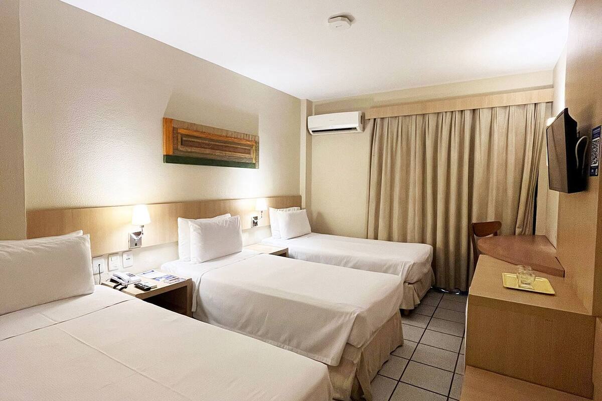 Quarto de hotel com duas camas, uma mesa, uma cadeira, televisão de tela plana e uma janela com cortinas.