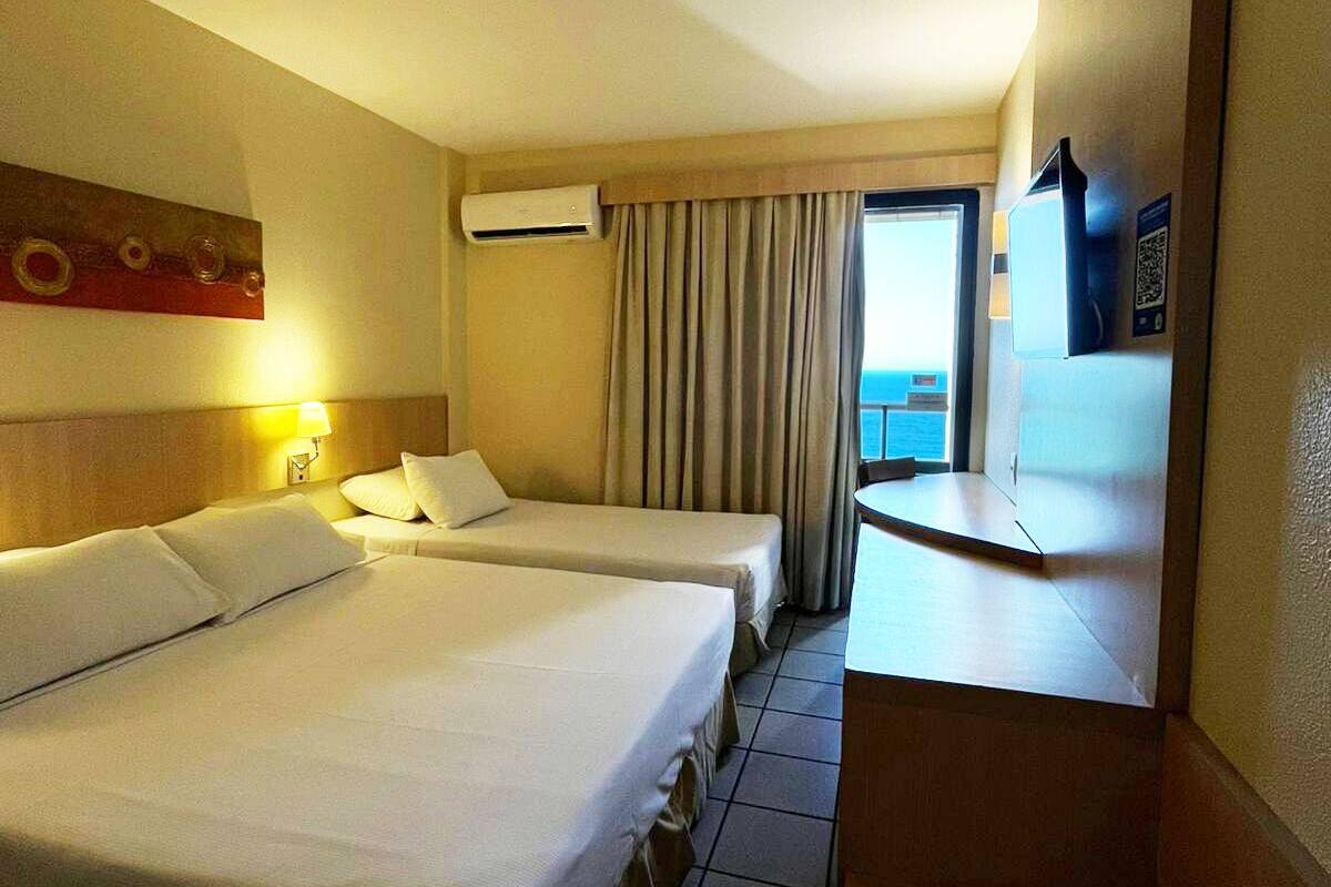 Quarto de hotel com duas camas, uma escrivaninha, aparelho de ar condicionado e vista para o mar.