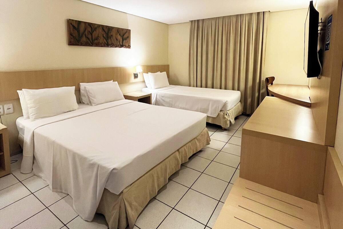 Quarto de hotel com duas camas, uma escrivaninha, televisão e um quadro na parede.