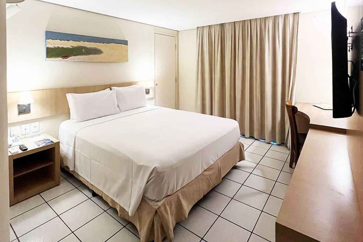 Quarto de hotel com uma cama grande, uma escrivaninha com cadeira, televisão e um quadro na parede.
