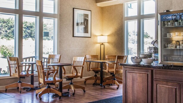 Uma sala de jantar com cadeiras e mesas de madeira, janelas amplas e uma área de cozinha com geladeira e prateleiras.