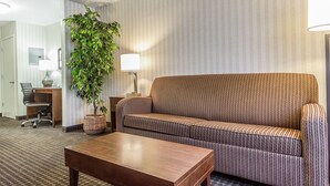 Quarto de hotel com sofá listrado, mesa de centro de madeira, escrivaninha com cadeira, luminária e planta em vaso.
