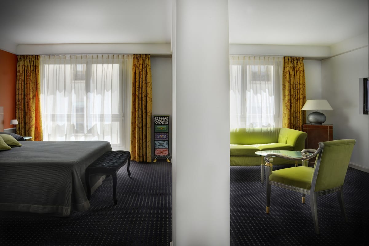 Quarto de hotel com uma cama, uma poltrona verde, uma mesa pequena e um abajur.