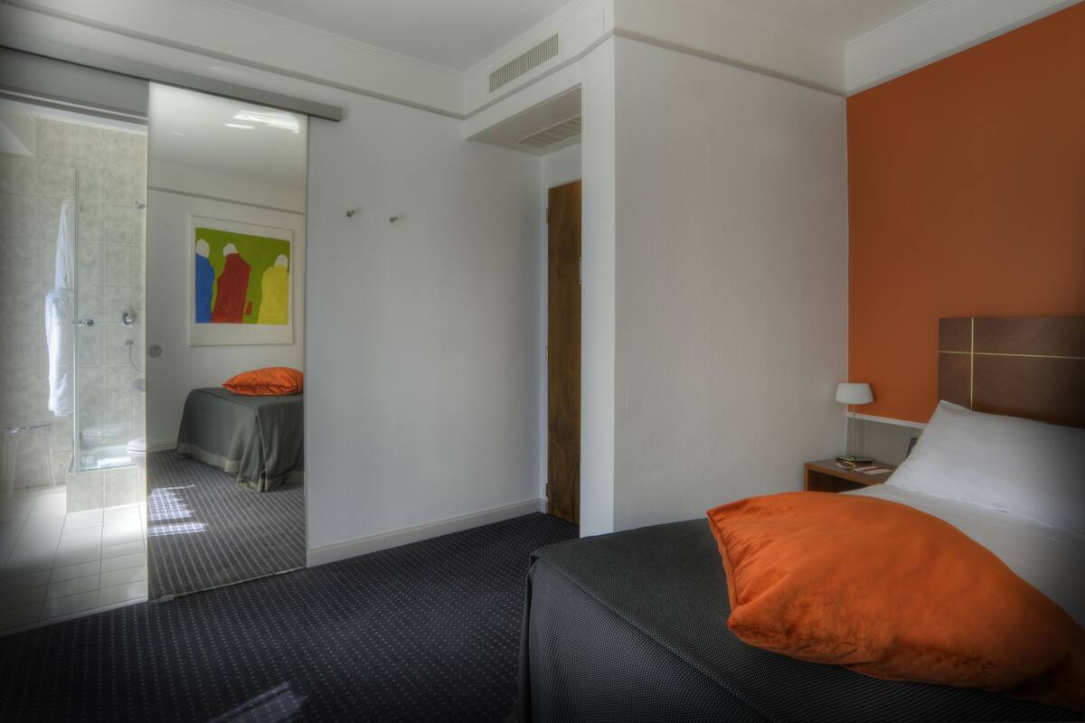 Quarto de hotel com cama, mesa de cabeceira, luminária, banheiro com chuveiro e porta que dá para outro quarto.