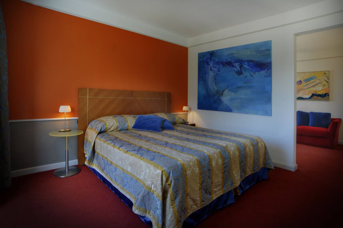 Quarto de hotel com uma cama grande, duas mesas de cabeceira, um tapete vermelho e um grande quadro abstrato na parede.