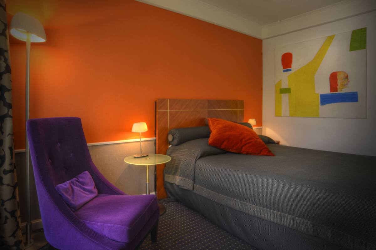 Quarto de hotel com uma cama, uma poltrona roxa, uma mesa redonda pequena e uma obra de arte na parede.