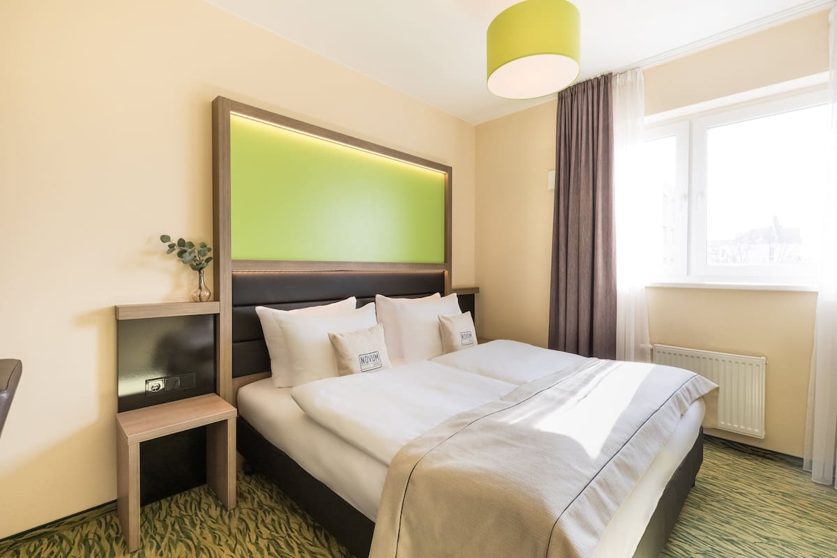 Quarto de hotel com uma cama grande, mesa de cabeceira, luminária e janela com cortinas.