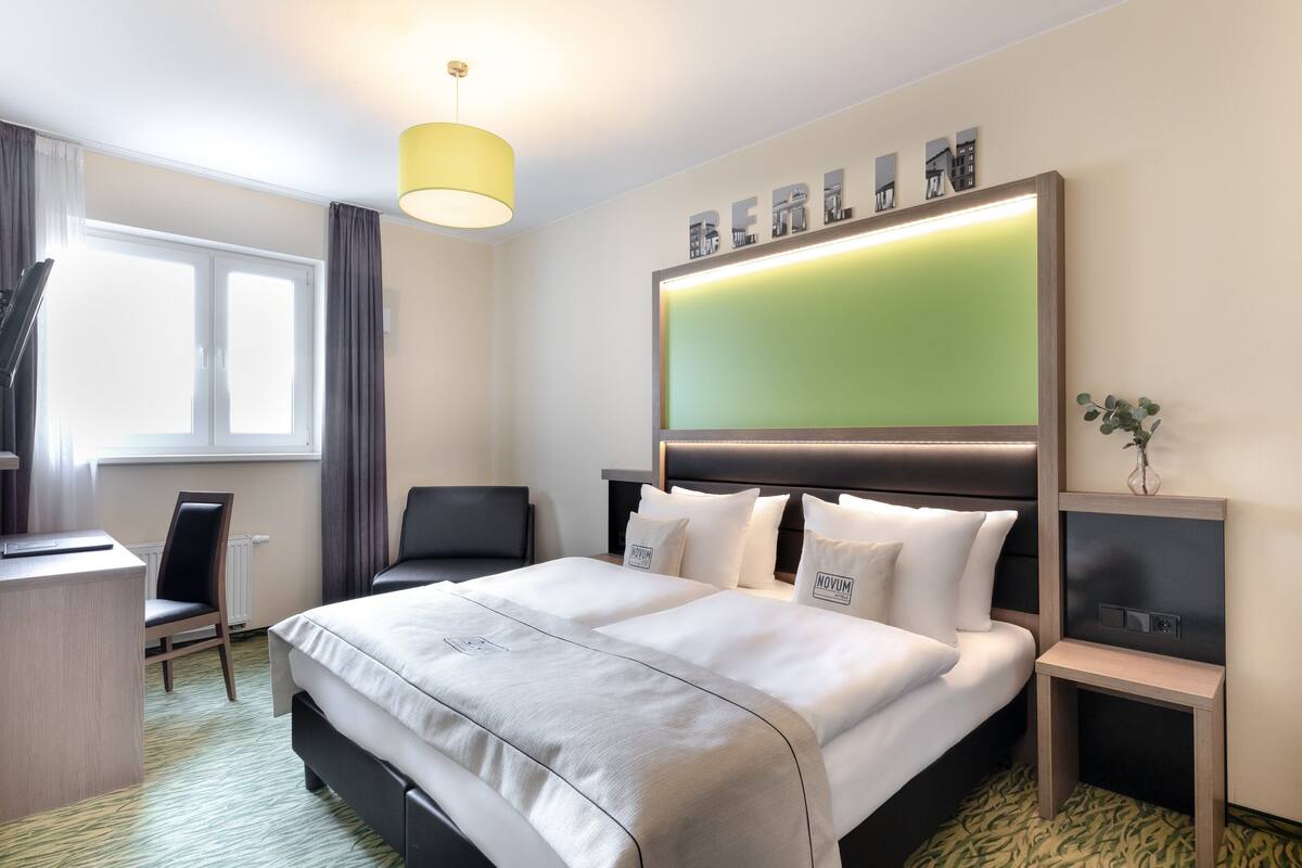 Quarto de hotel com uma cama grande, uma escrivaninha, uma cadeira e uma mesinha de cabeceira.