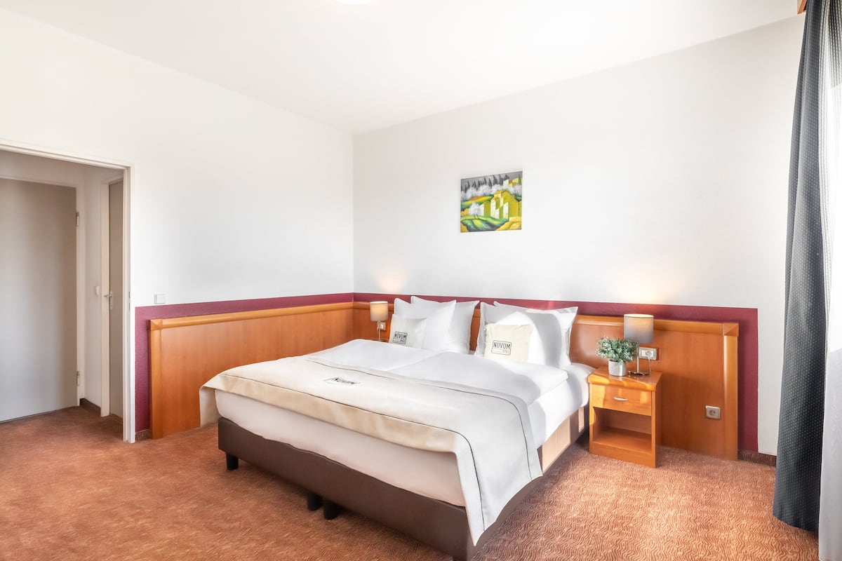 Quarto de hotel com uma cama grande, cabeceira de madeira, mesa de cabeceira com abajur, um quadro na parede e uma porta à esquerda.