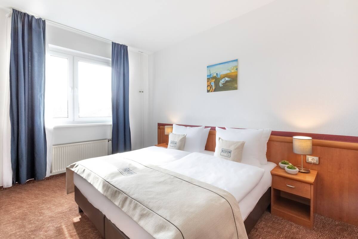 Quarto de hotel com uma cama grande, uma mesa de cabeceira com abajur e um quadro na parede.