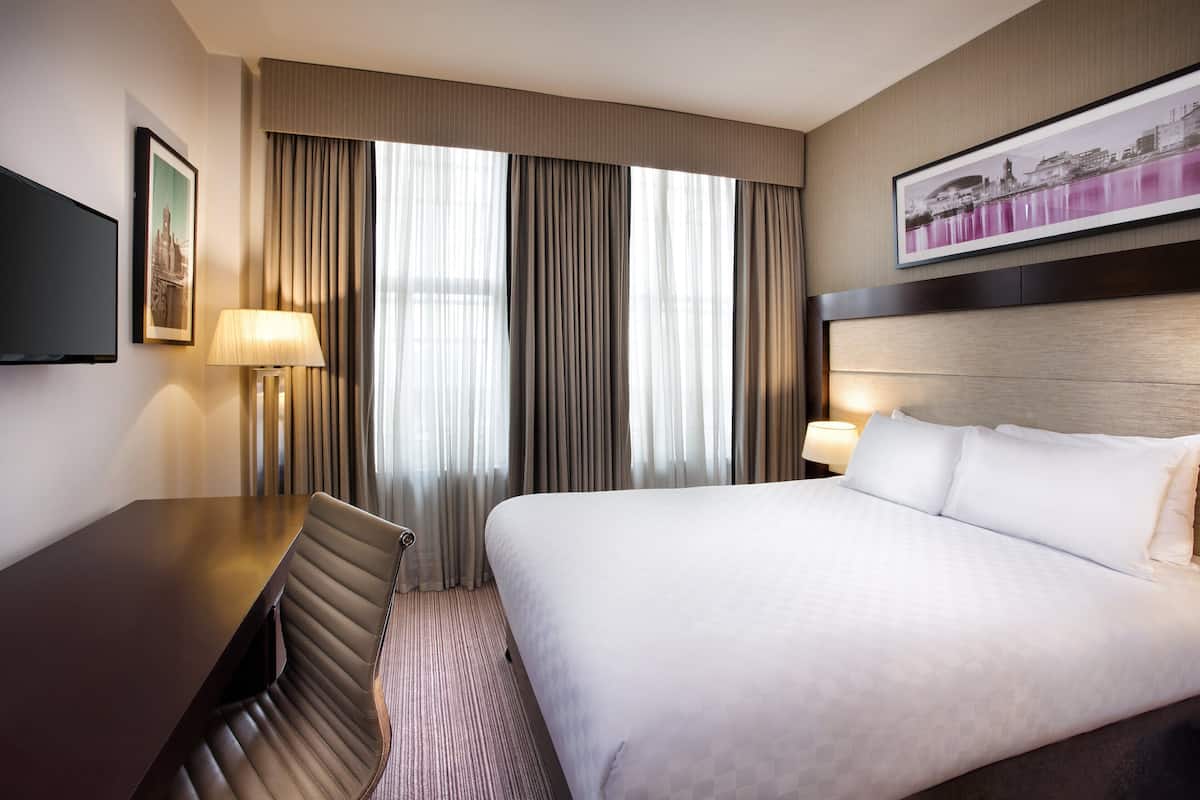Quarto de hotel com cama, escrivaninha, cadeira, televisão e janela com cortinas.