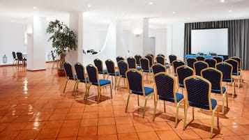Uma sala de conferências com fileiras de cadeiras, uma tela grande e um vaso de plantas.
