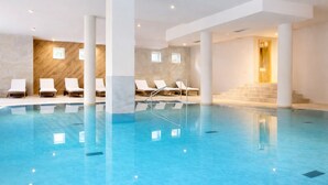 Piscina interna com cadeiras de descanso brancas e parede de mármore.