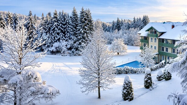 Paisagem coberta de neve com um edifício com varandas, piscina e uma floresta ao fundo.