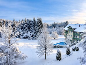 Paisagem coberta de neve com um edifício com varandas, piscina e uma floresta ao fundo.