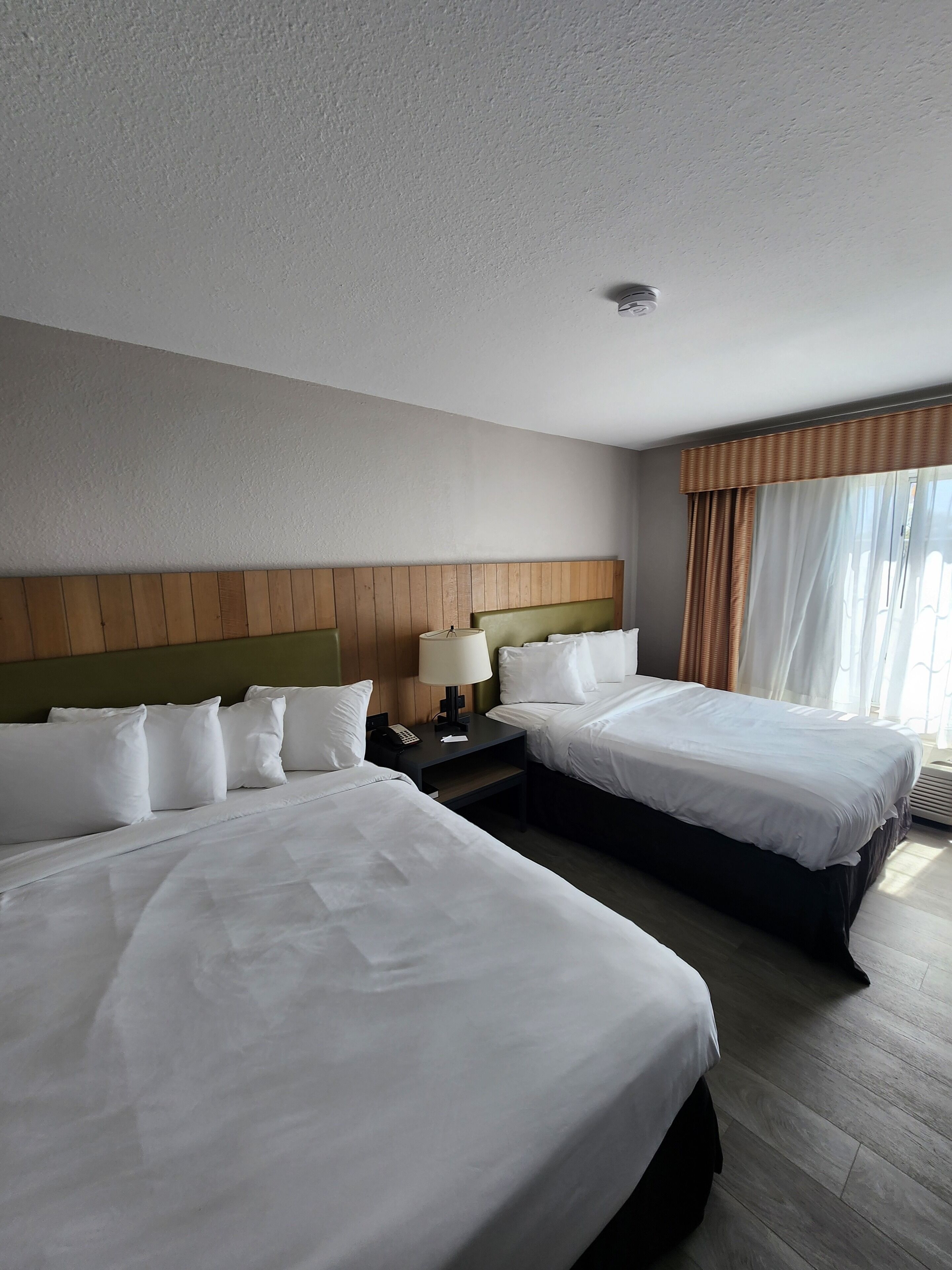 Quarto de hotel com duas camas, uma mesa de cabeceira com abajur e uma janela com cortinas.