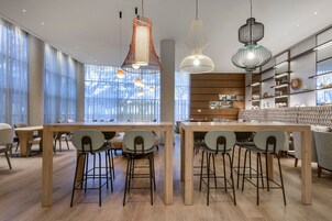 Uma área de jantar moderna com mesas de madeira e banquetas, janelas amplas com cortinas leves e iluminação pendente.
