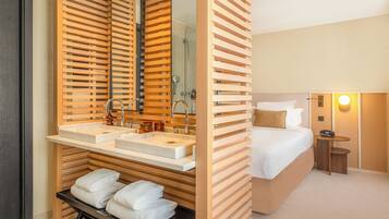 Quarto de hotel com parede de ripas de madeira, bancada com duas pias, cama com cabeceira, mesa de cabeceira e criado-mudo.