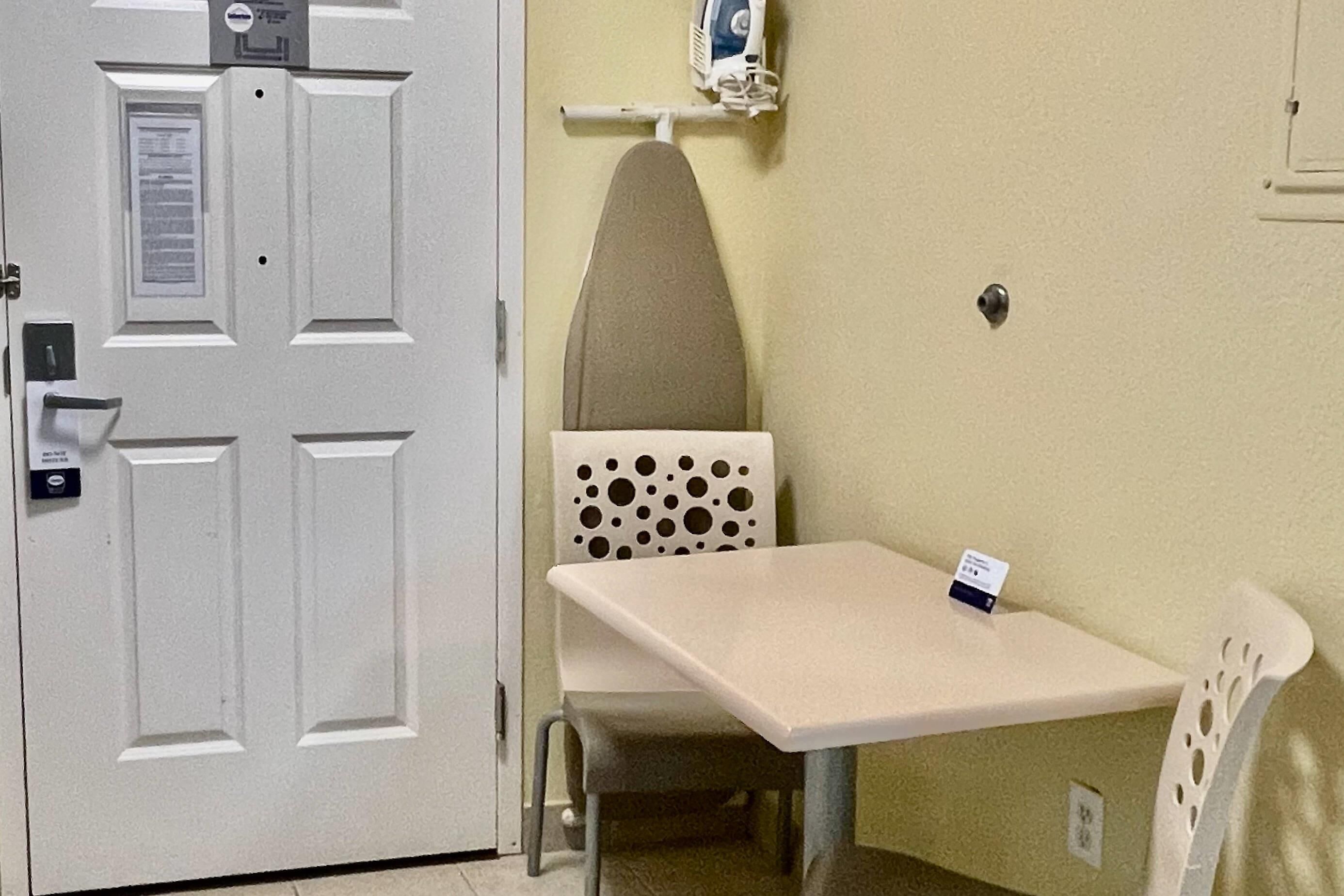 Um quarto pequeno com uma porta branca, uma mesa e uma cadeira. Há uma pequena placa na mesa e um dispositivo fixado na parede acima da cadeira.