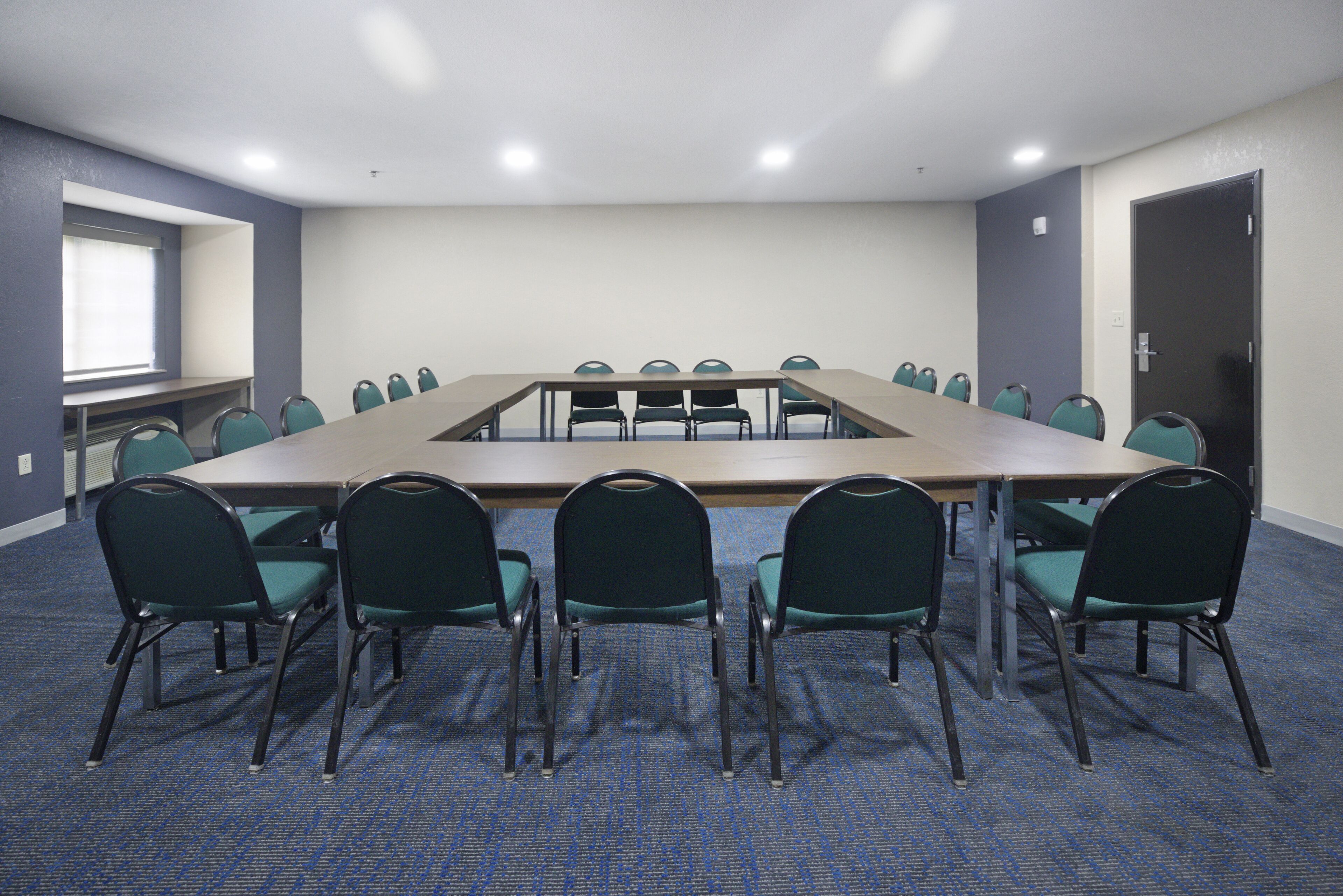 Sala de conferências com uma mesa longa e cadeiras, piso com carpete e tela de projetor.