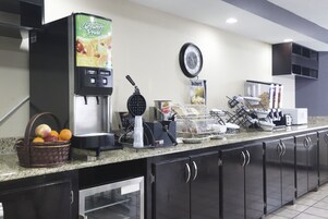 Uma área de cozinha com dispensador de suco, máquina de waffles e balcão de pastelaria.