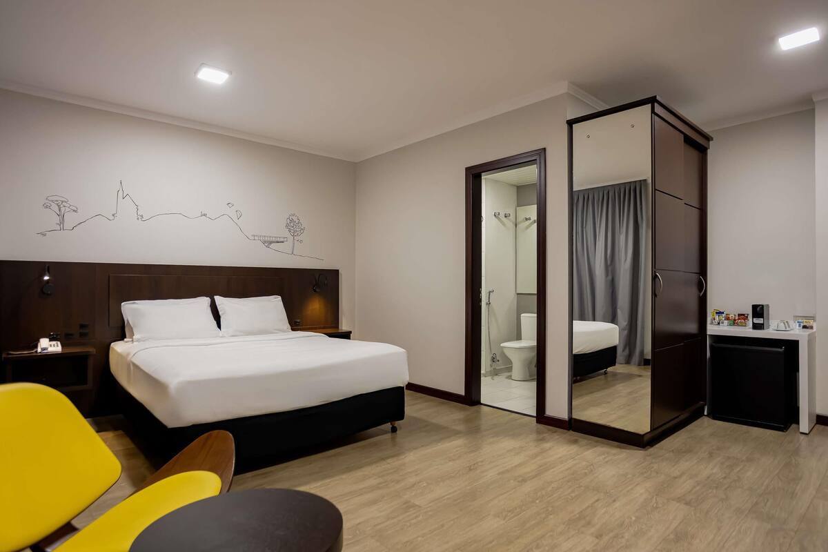 Quarto de hotel com cama, secretária, cadeira, espelho e área de chuveiro.