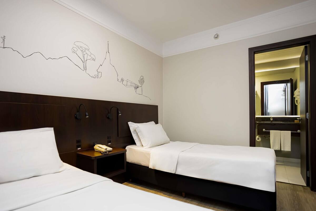 Quarto de hotel com duas camas, cabeceira de madeira, obra de arte na parede, mesa de cabeceira com luminária e banheiro visível por uma porta aberta.