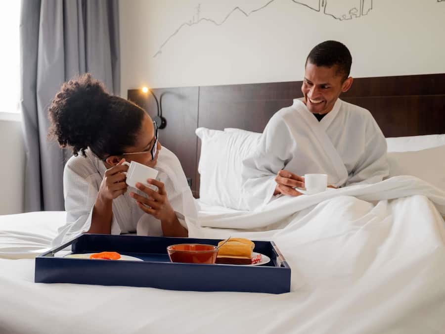 Quarto de hotel com uma cama, um tabuleiro com comida e bebidas, e uma parede com um desenho a linha.