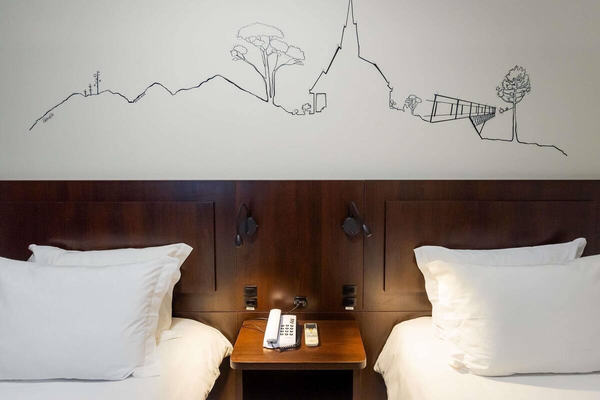 Quarto de hotel com duas camas, cabeceira de madeira, mesa de cabeceira com telefone e um mural na parede retratando uma paisagem.