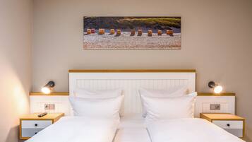 Uma cama de casal com lençóis brancos, duas mesas de cabeceira com abajures, um painel de cabeceira e uma pintura de cabanas de praia na parede.