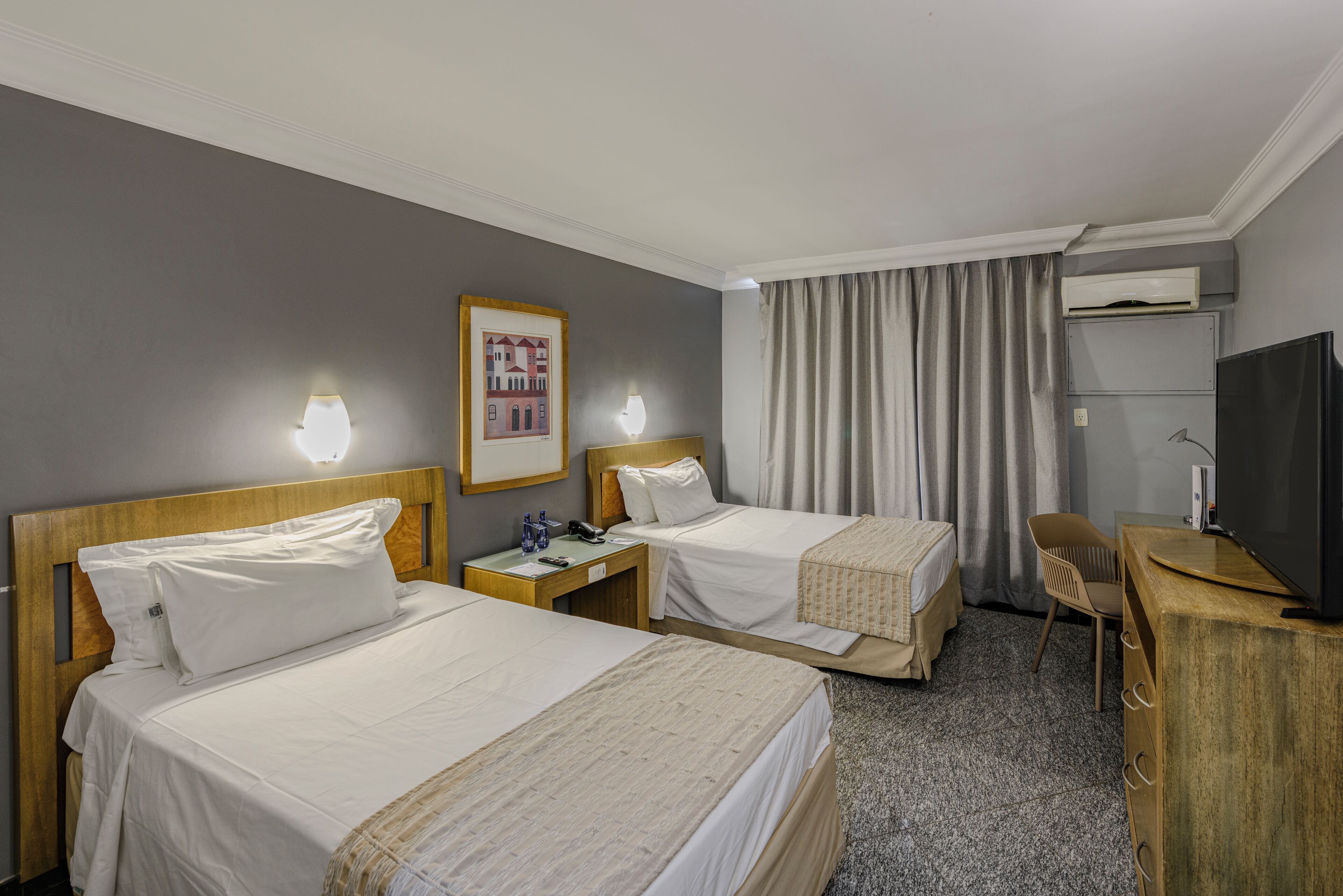 Quarto de hotel com duas camas, uma escrivaninha, uma cadeira, uma televisão e um quadro na parede.